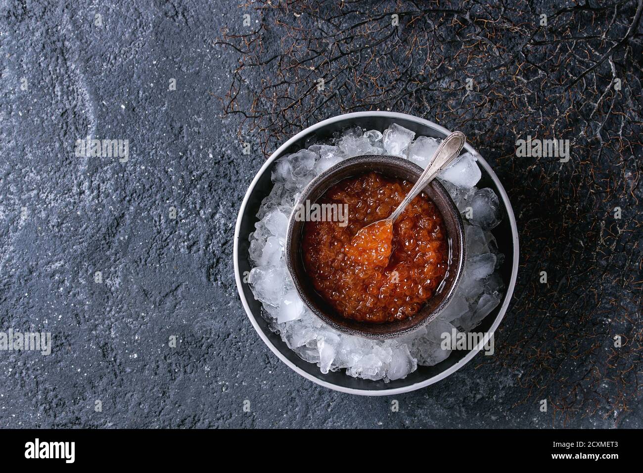 Bowl of red caviar on ice with spoon served over black stone texture ...
