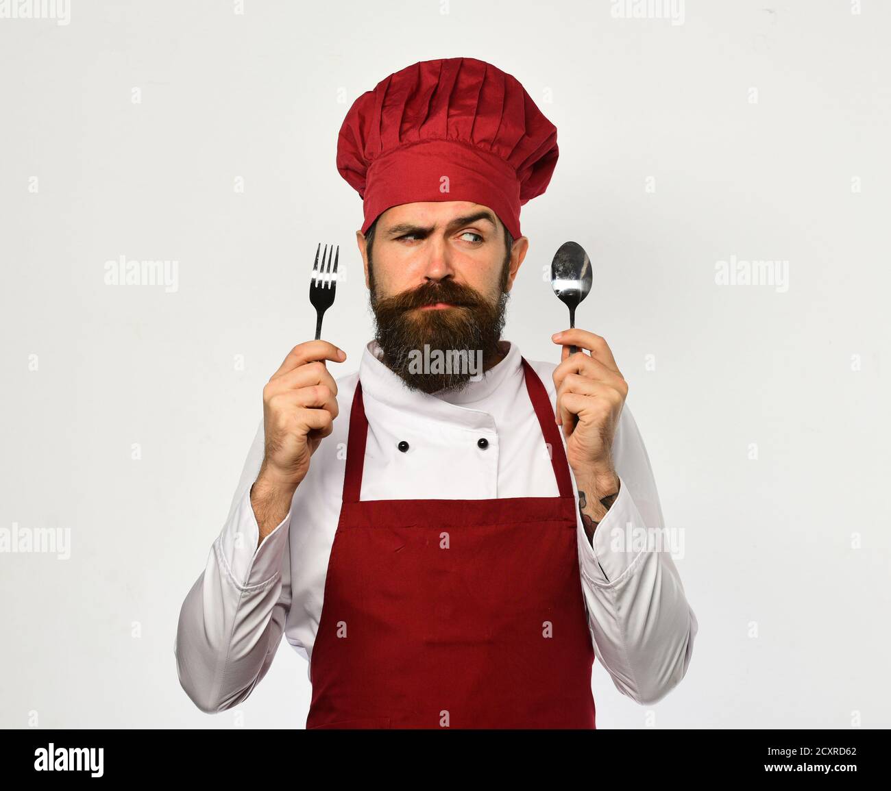 Cook with confused face in burgundy uniform shows cutlery. Chef ...
