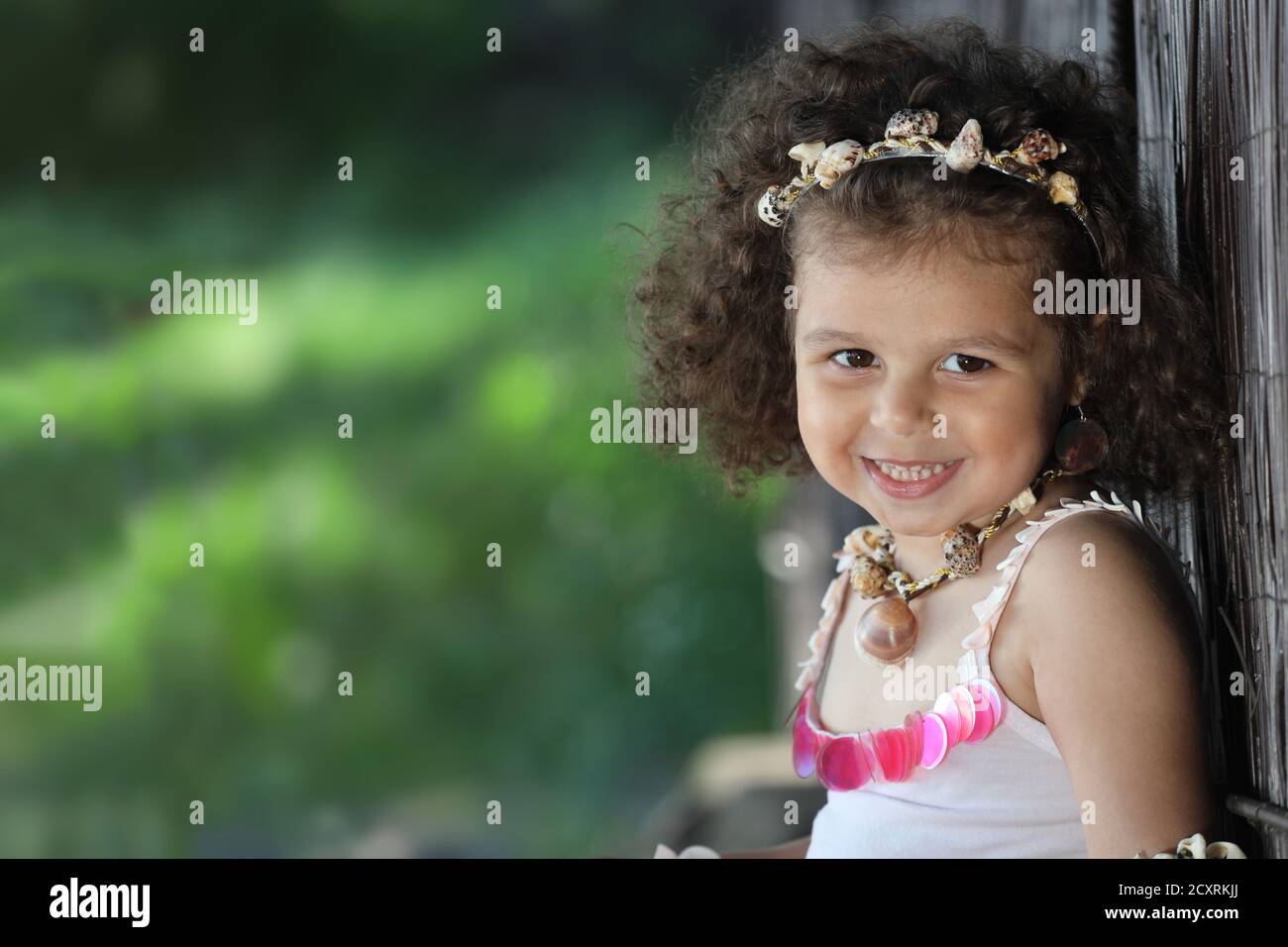 curly girl in a pink dress with shell decorations sits on the nature ...