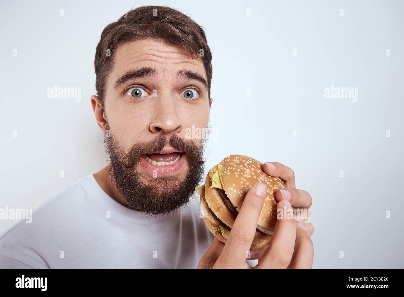 A man eating a hamburger on a light background in a white T-shirt ...