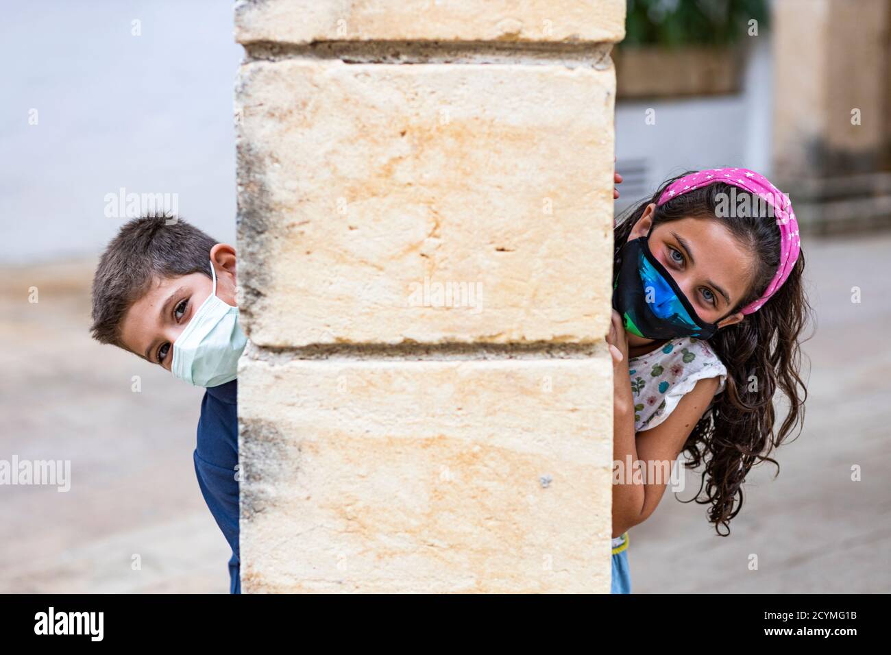 Two funny kids with face masks behind a column Stock Photo - Alamy