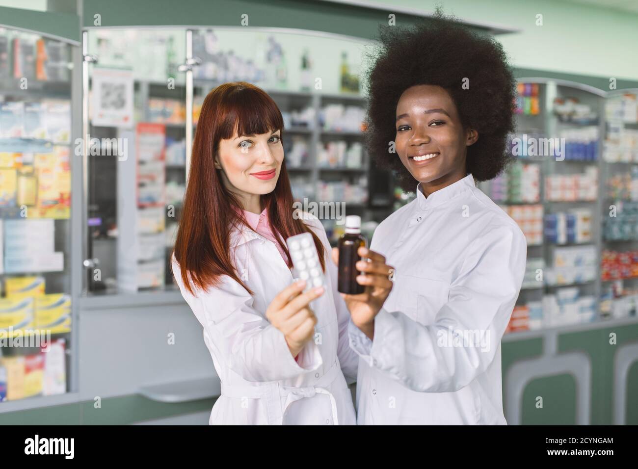 Team of two smiling multiethnical female chemists in pharmacy standing ...