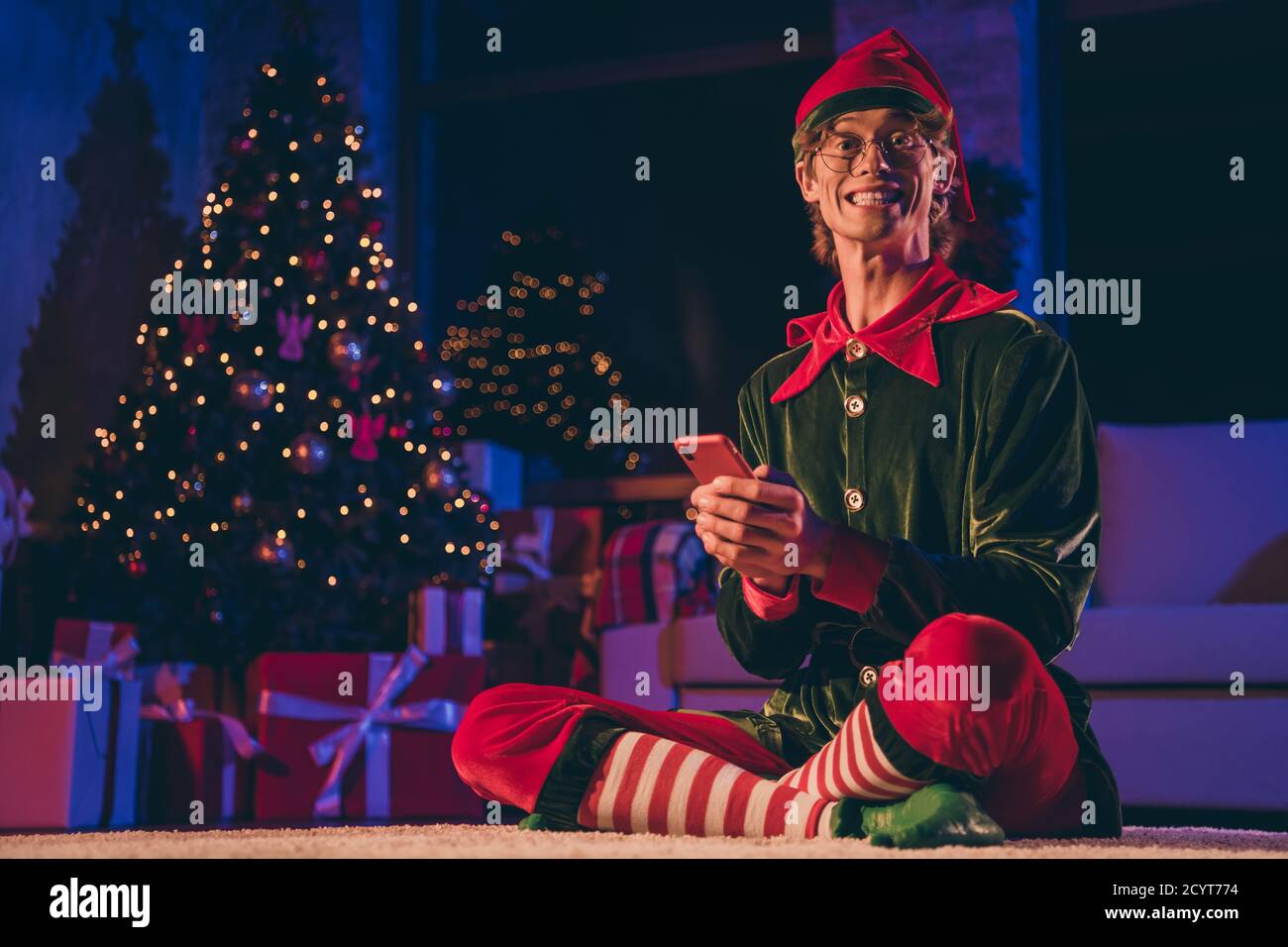 Full length photo of funky elf sit floor have smartphone wear green hat ...