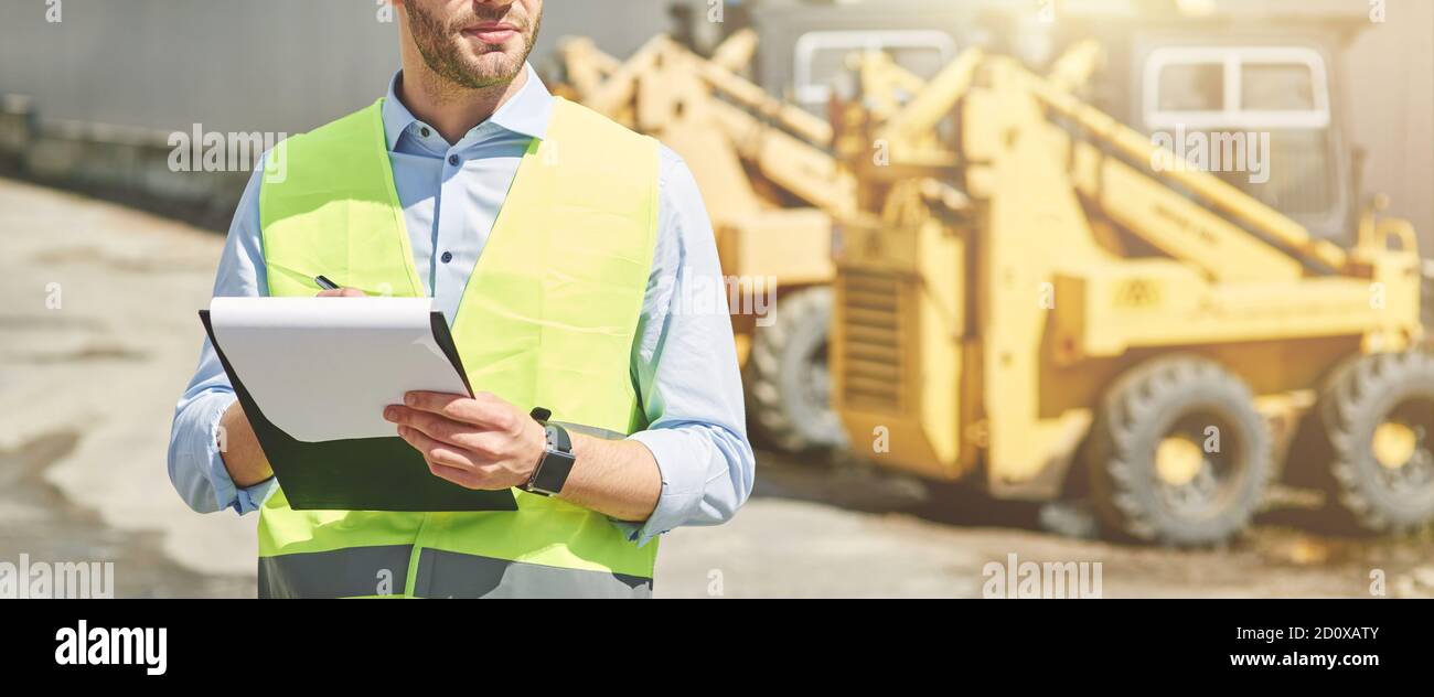 Young civil engineer wearing helmet writing report while standing at ...