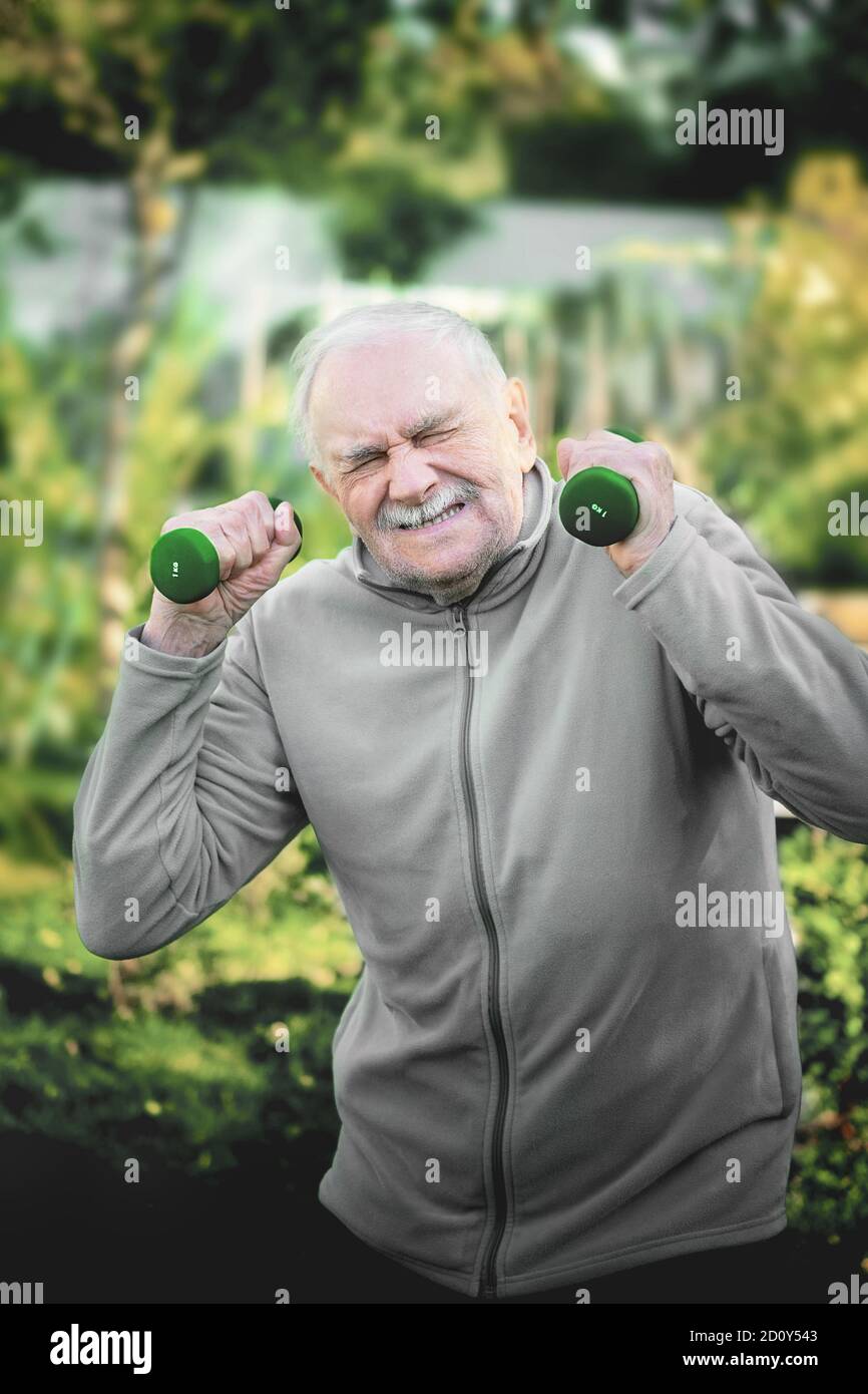 An old man trains his muscles with dumbbells in the Park after a home ...