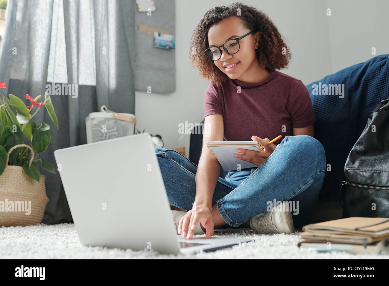 Happy young cross-legged student studying on the floor by bed in front ...
