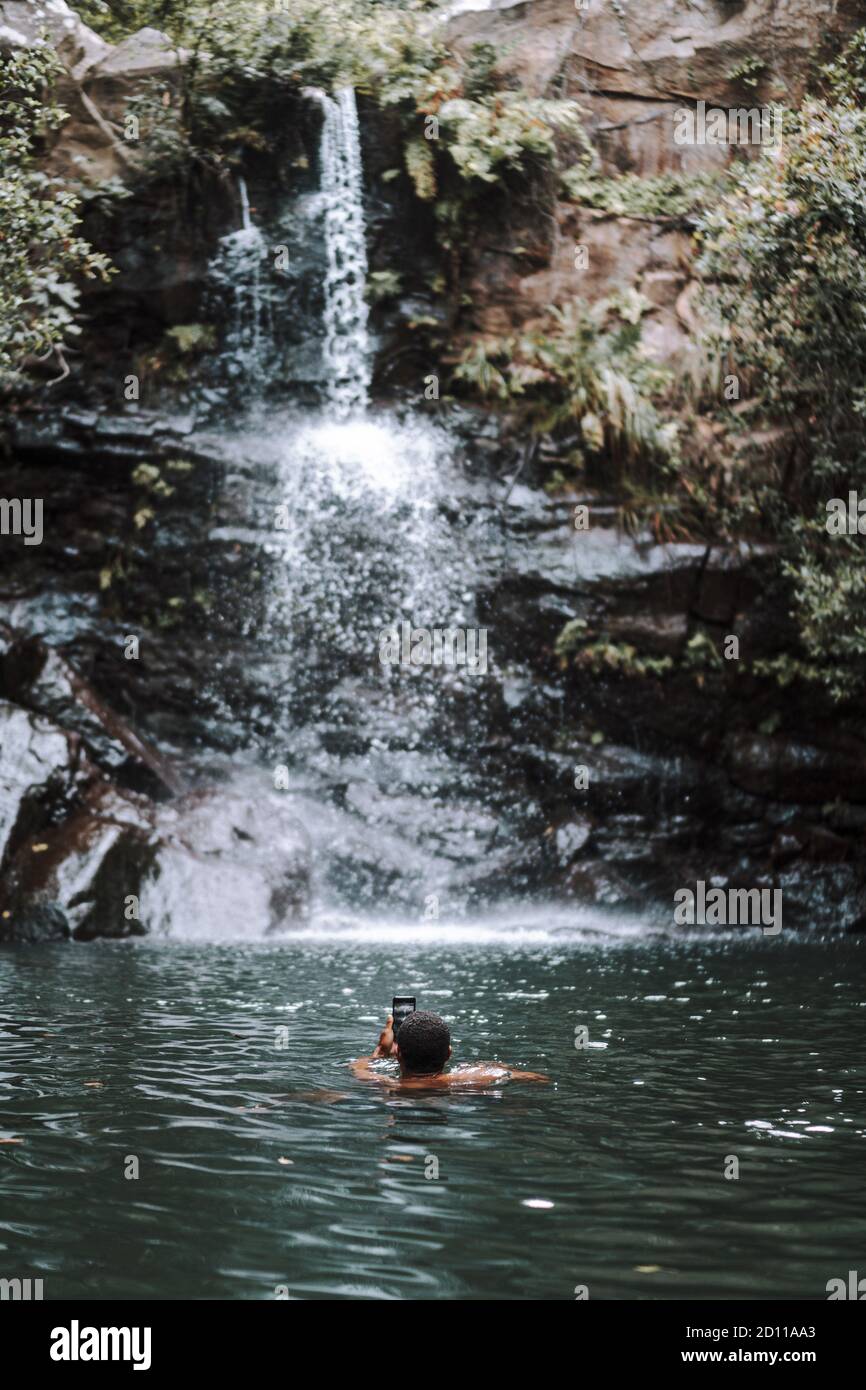 Vertical shot of a male taking a picture of a beautiful waterfall while ...