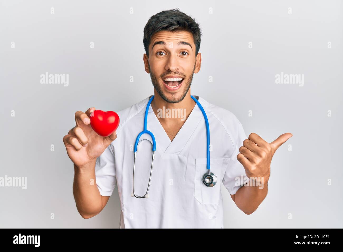 Young handsome man wearing doctor uniform holding heart pointing thumb ...