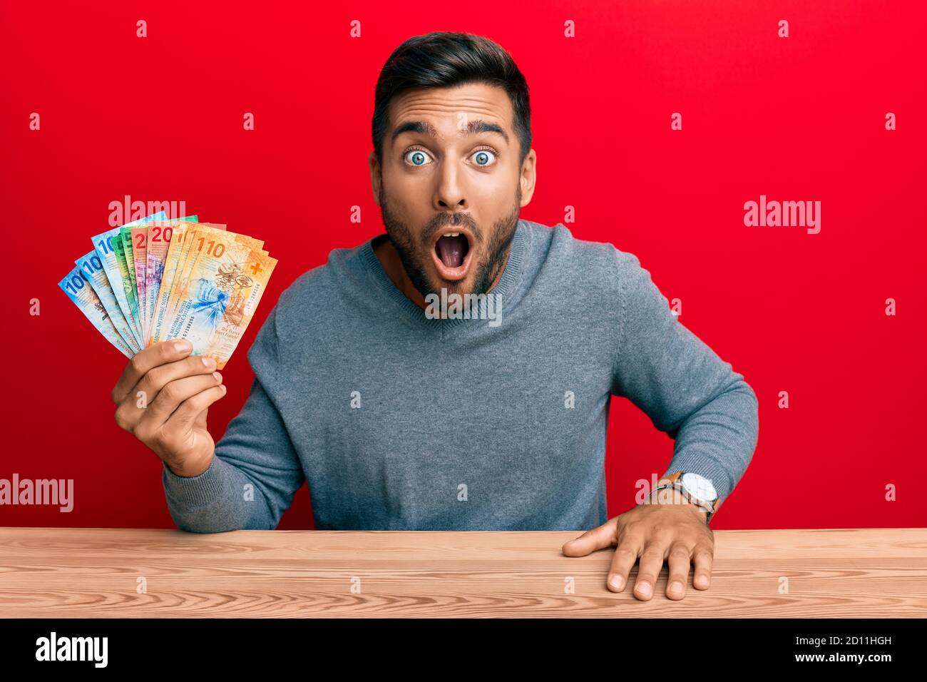 Handsome hispanic man holding swiss franc banknotes scared and amazed ...