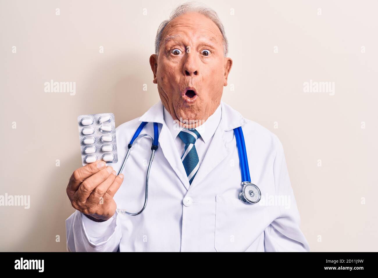 Senior grey-haired doctor man wearing stethoscope holding pills over ...
