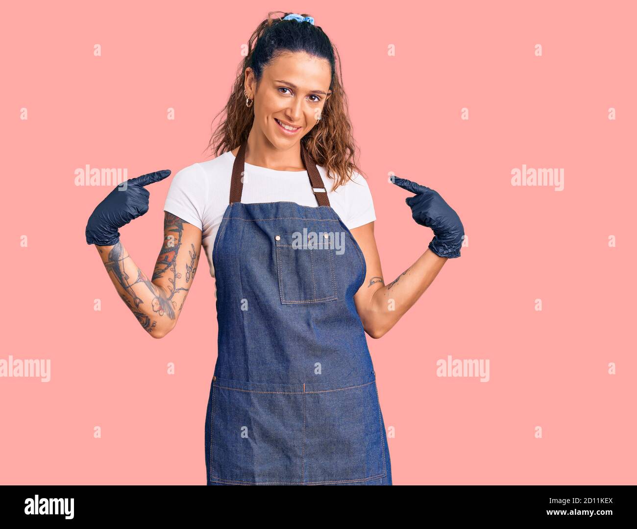 Young hispanic woman with tattoo wearing barber apron and gloves ...