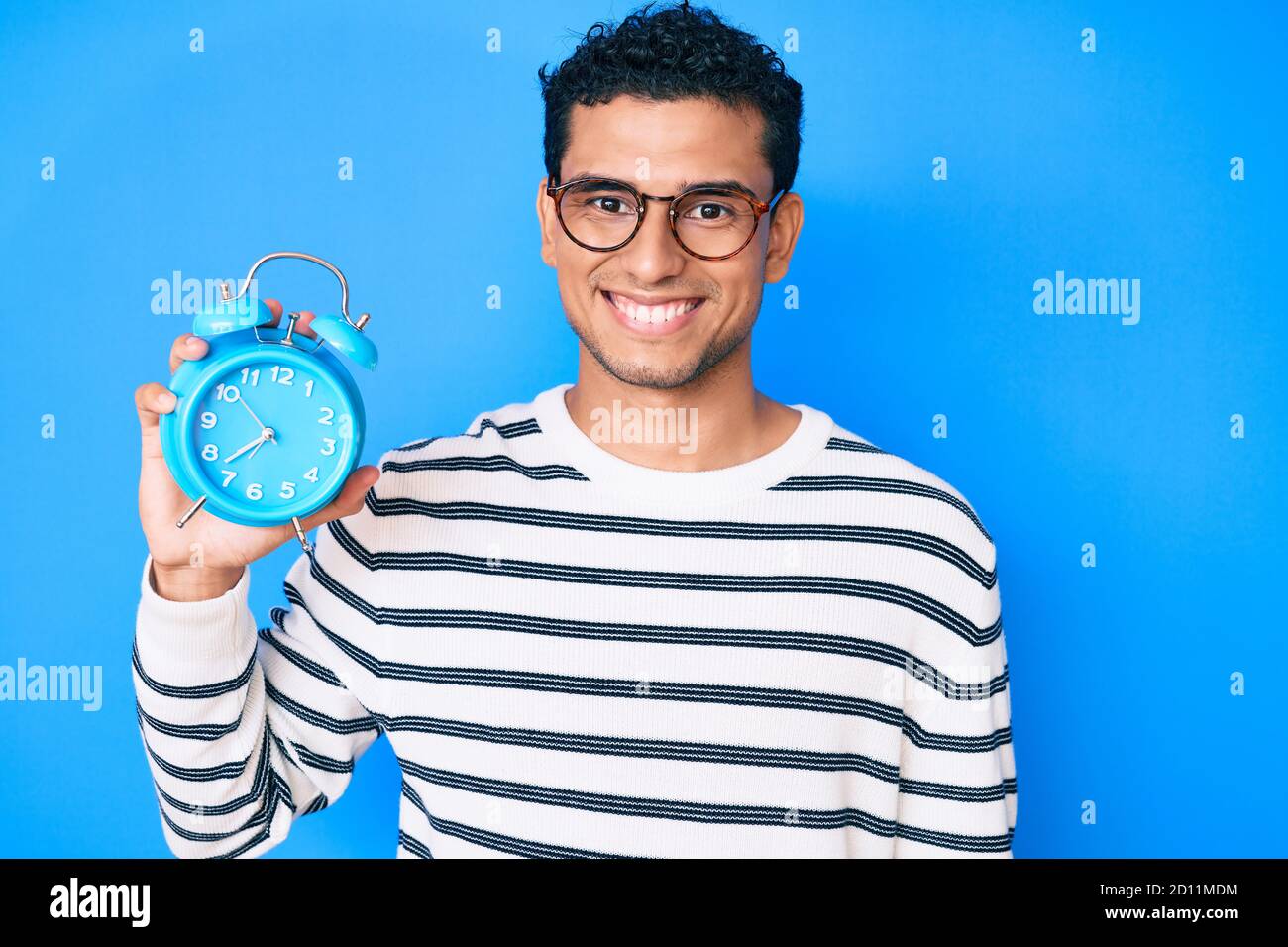 Young handsome hispanic man holding alarm clock looking positive and ...