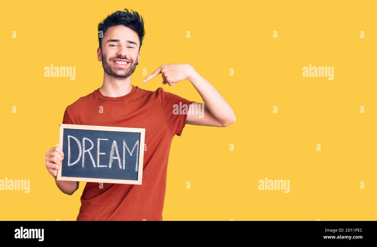 Young hispanic man holding blackboard with dream word pointing finger ...