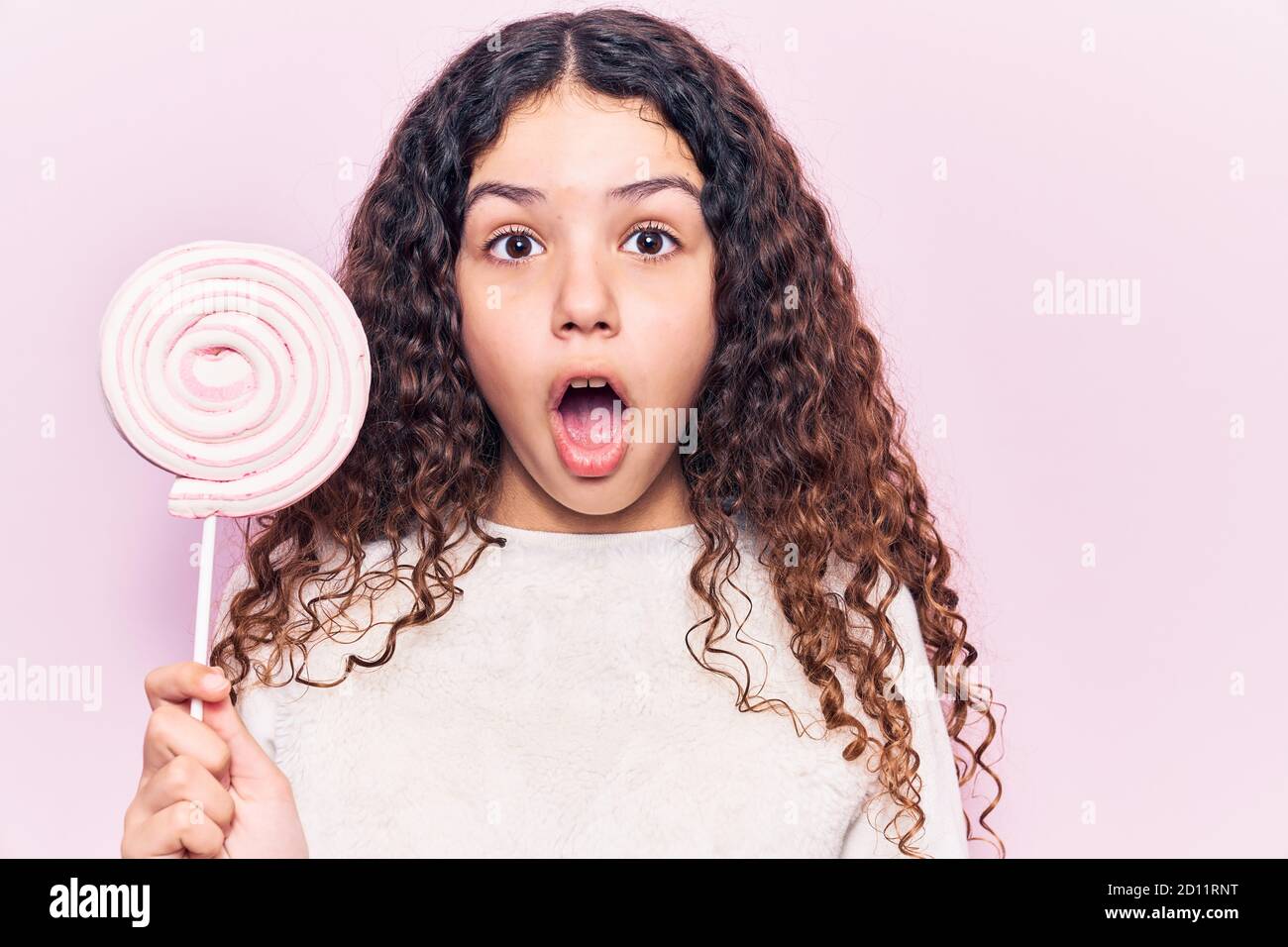 Beautiful kid girl with curly hair holding lollipop scared and amazed ...