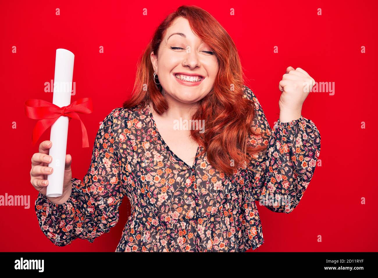 Young beautiful redhead woman holding graduated degree diploma over red ...