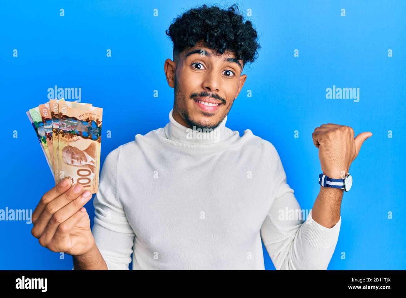 Young arab handsome man holding canadian dollars pointing thumb up to ...