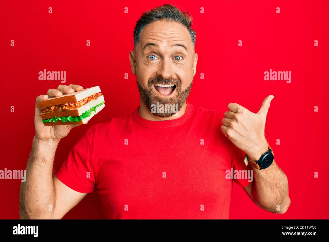 Handsome middle age man eating sandwich pointing thumb up to the side ...