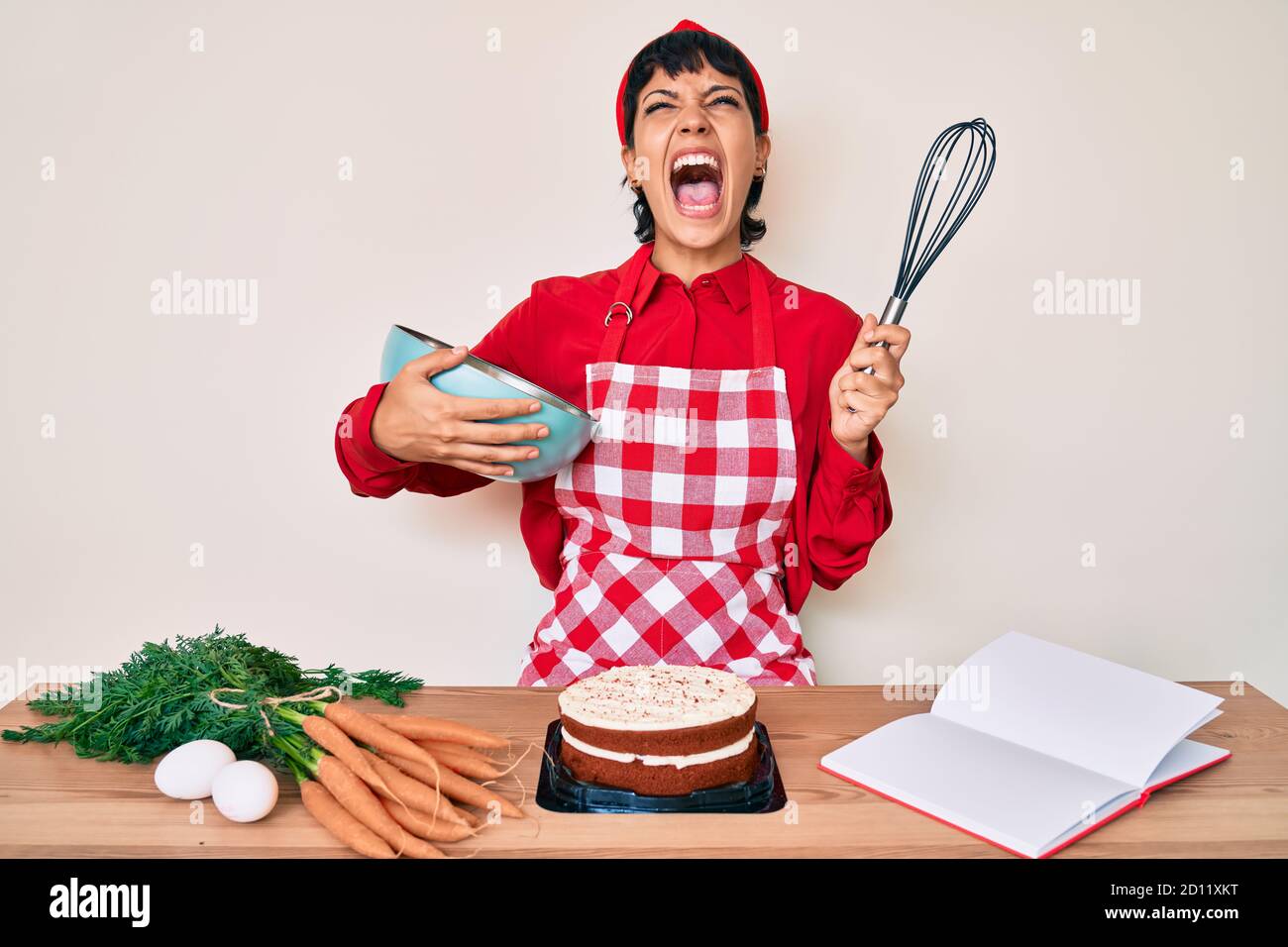 Beautiful brunettte woman cooking carrot cake using whisk angry and mad ...