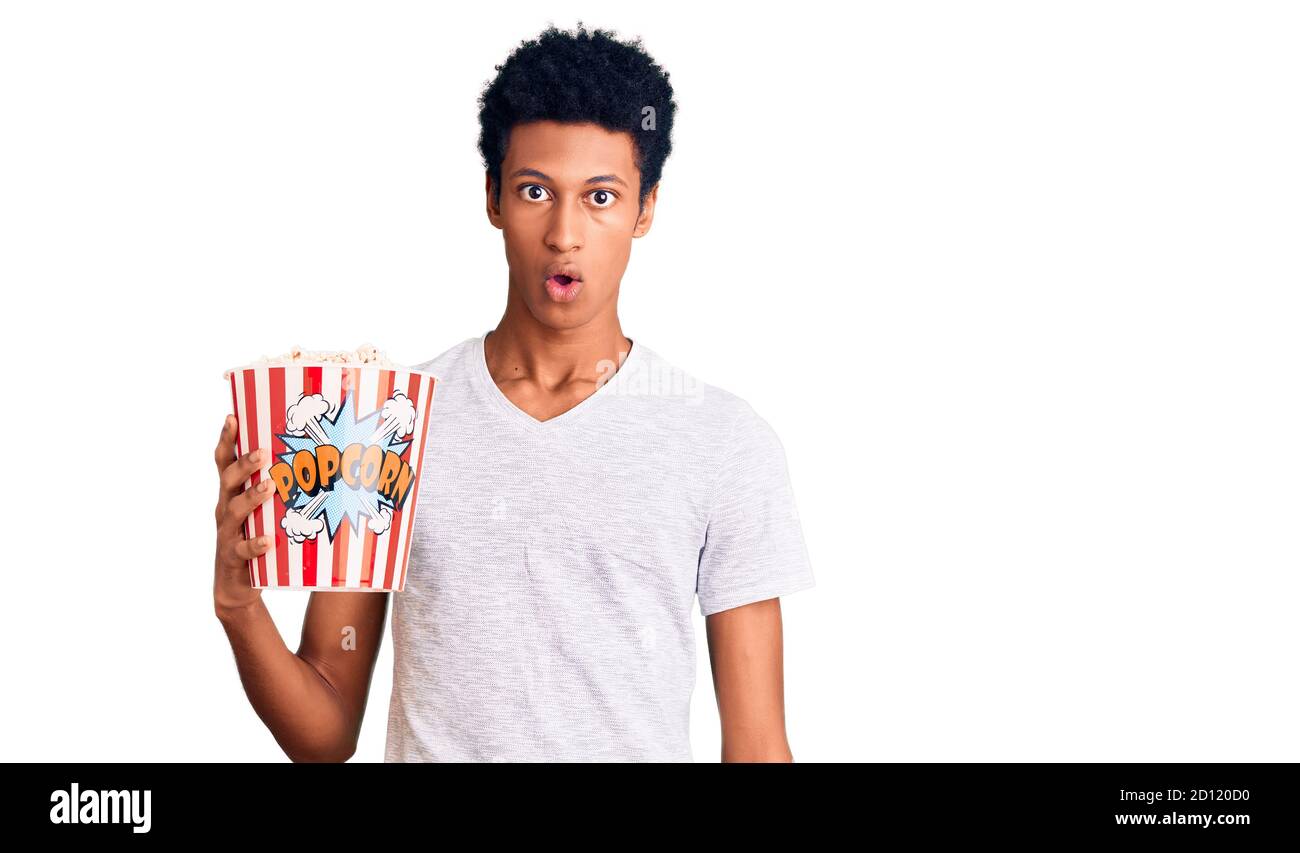 Young african american man holding popcorn scared and amazed with open mouth for surprise ...