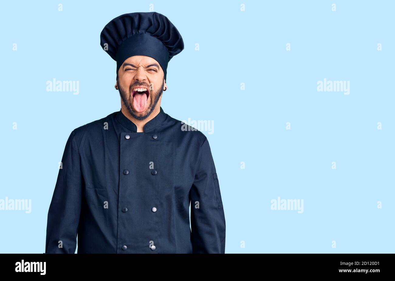 Young hispanic man wearing cooker uniform sticking tongue out happy ...