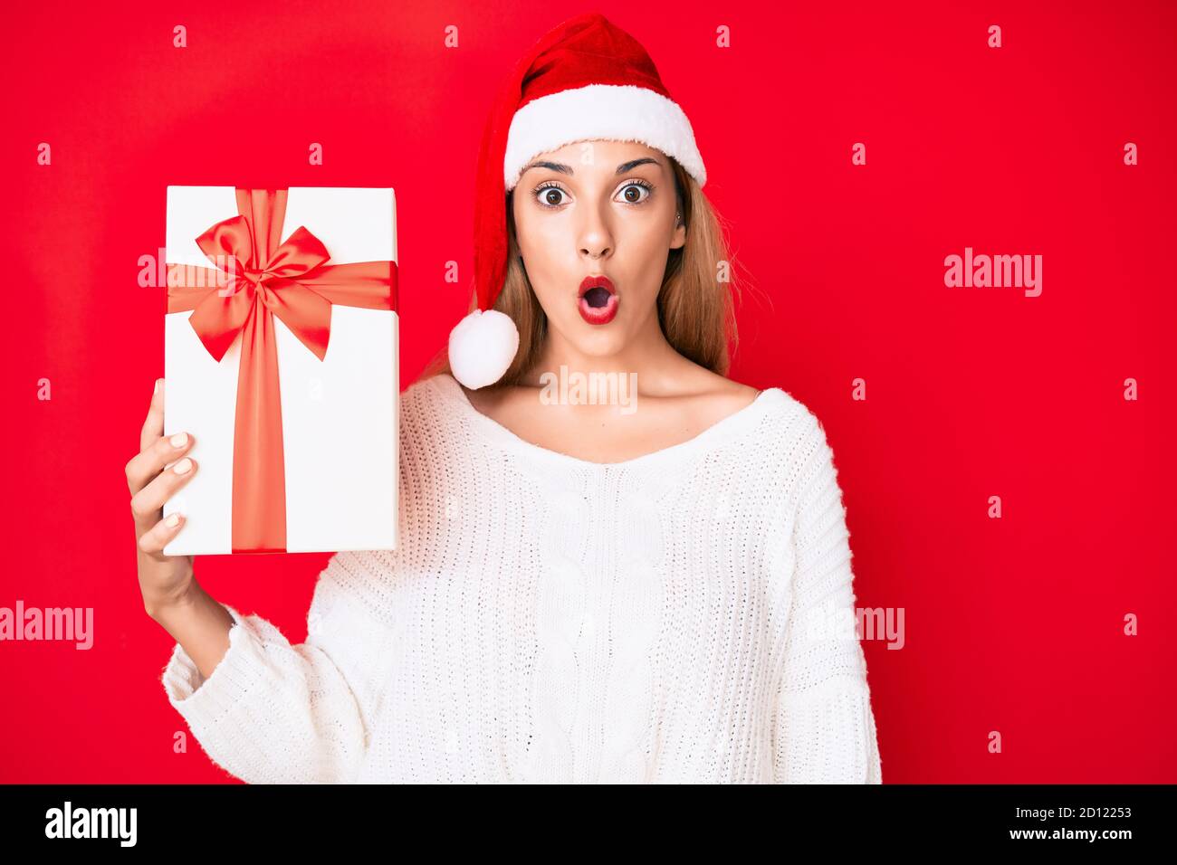 Young brunette woman wearing christmas hat and holding a gift scared ...