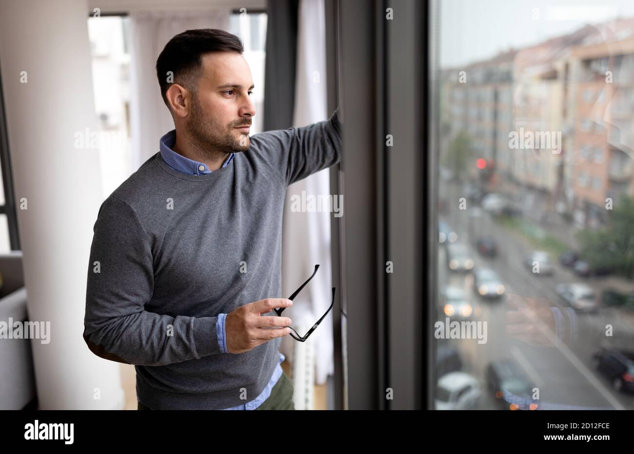 Portrait of man sad and depressed standing at window in despair at home ...