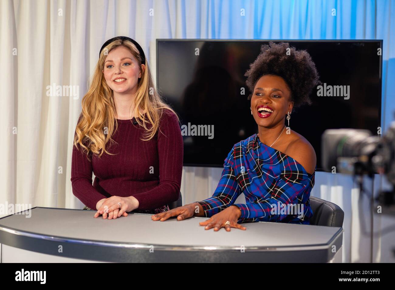 Two TV hosts sitting at a desk in a film studio, filming a TV show ...