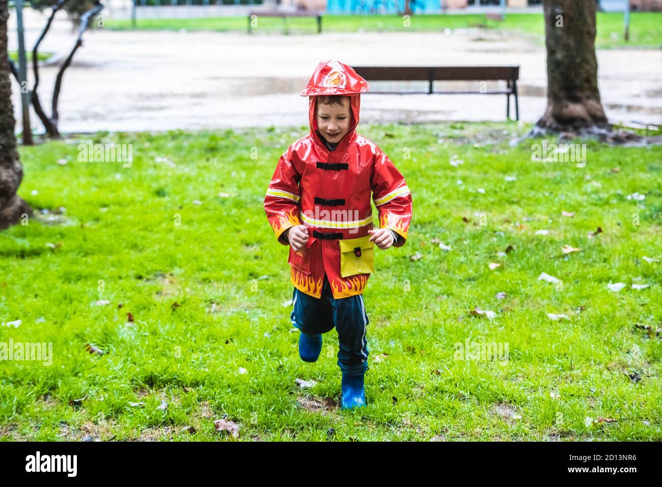 Boy dressed as a fireman with red raincoat splashes on the grass of a ...