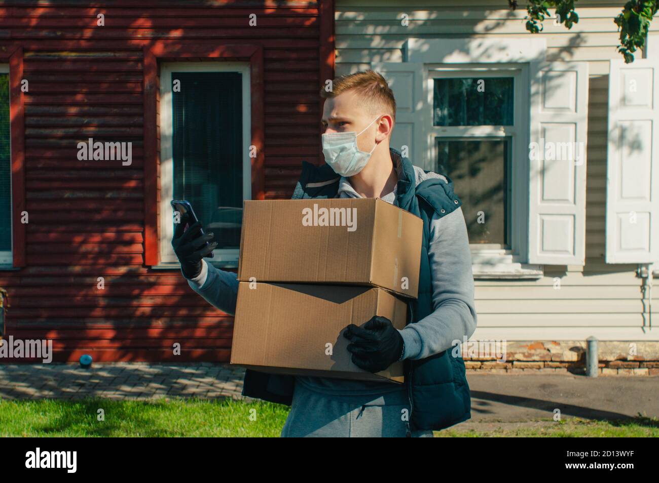 Delivery man wearing a mask and gloves holds two boxes and looks at his ...