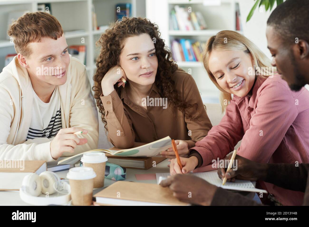 Multi-ethnic group of smiling young people studying together while sitting at table in college ...