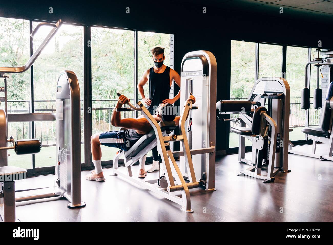 trainer wearing a face mask helps a young man do his workout at the gym ...