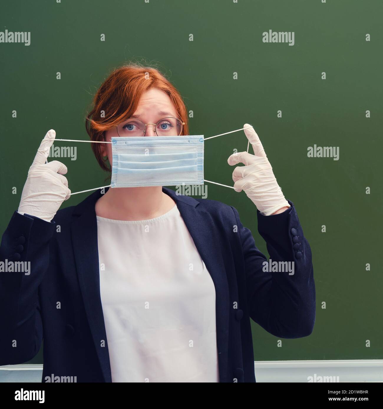 Woman teacher next to the school blackboard puts on a protective mask ...