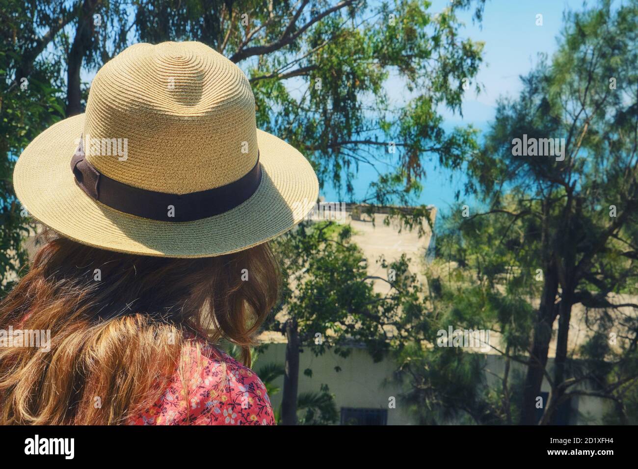 A woman in a straw hat looks at the sea behind the branches of trees in ...