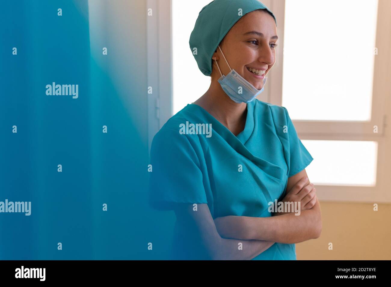 Positive young medical worker in uniform with mask standing near window ...