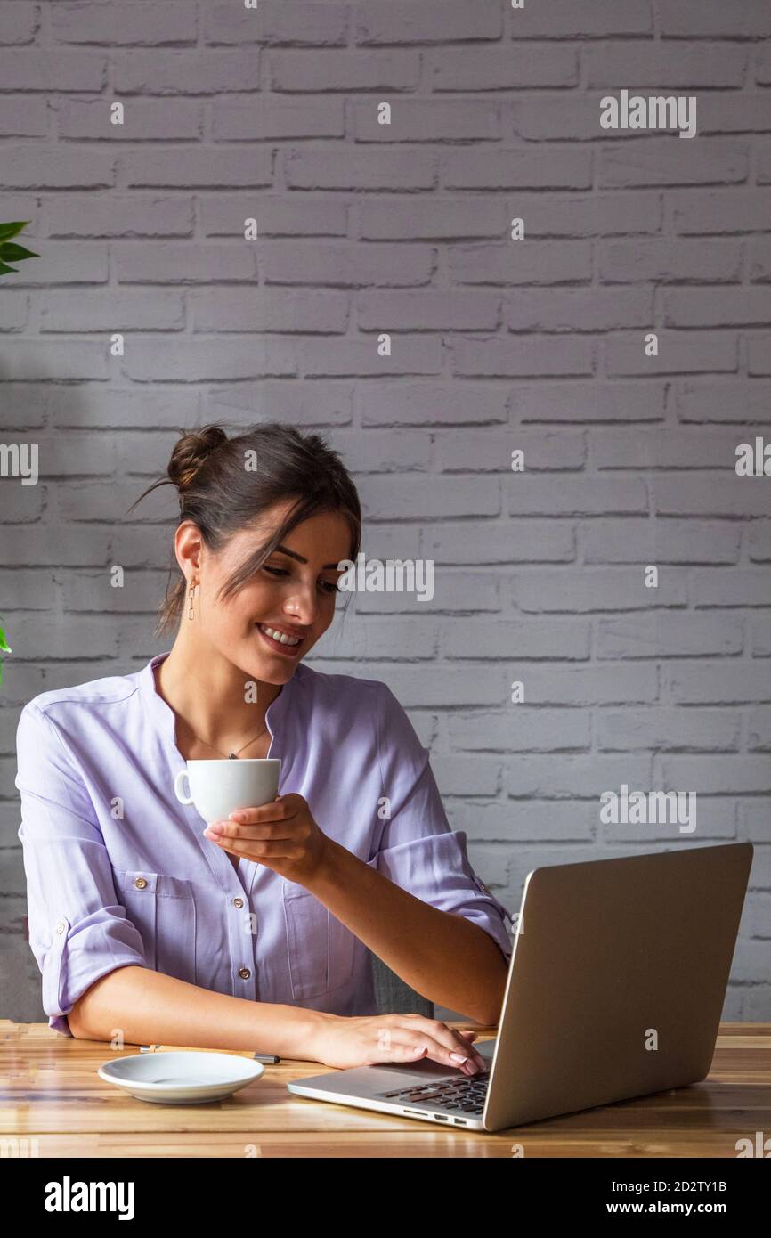 Smiling young female remote employee with cup of coffee in hand sitting ...