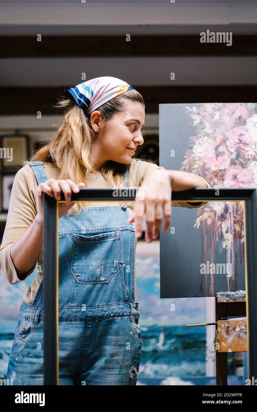 Creative young female artist in denim overall leaning on empty frame in ...