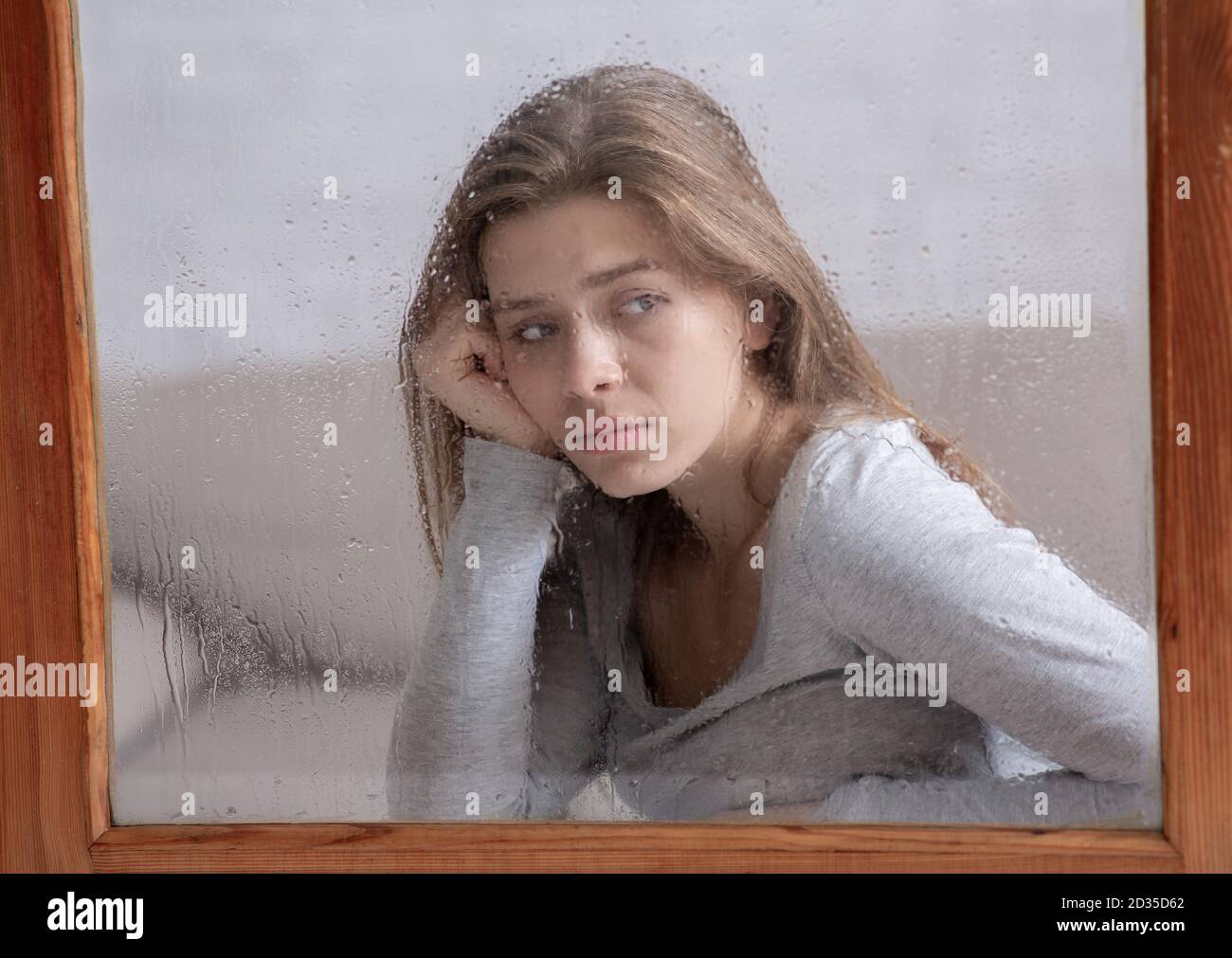 Sad young woman sitting at home and looking out window at falling rain ...