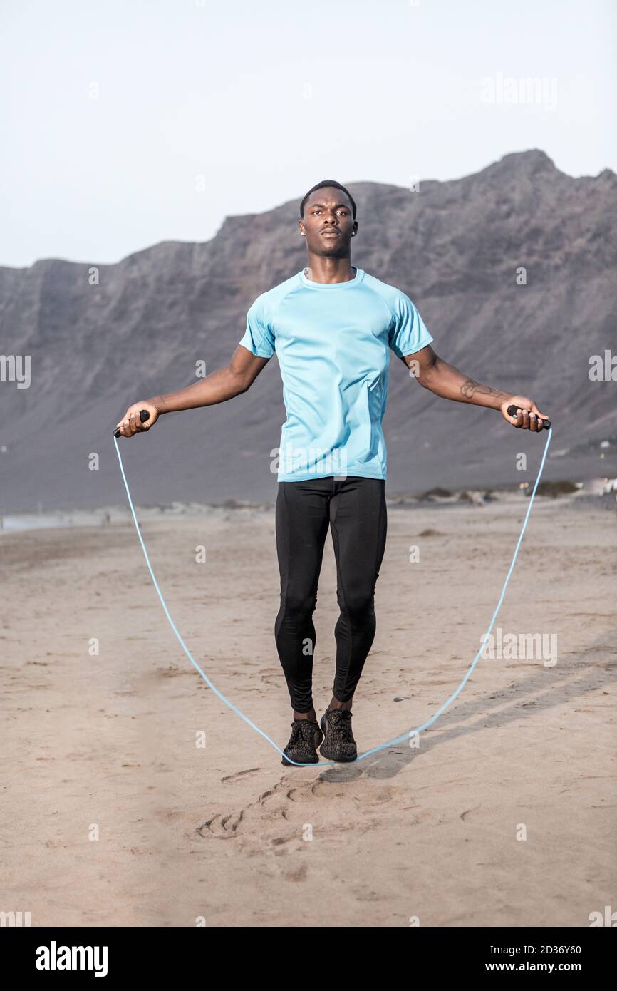 Full length African American male athlete jumping with rope on sandy ...