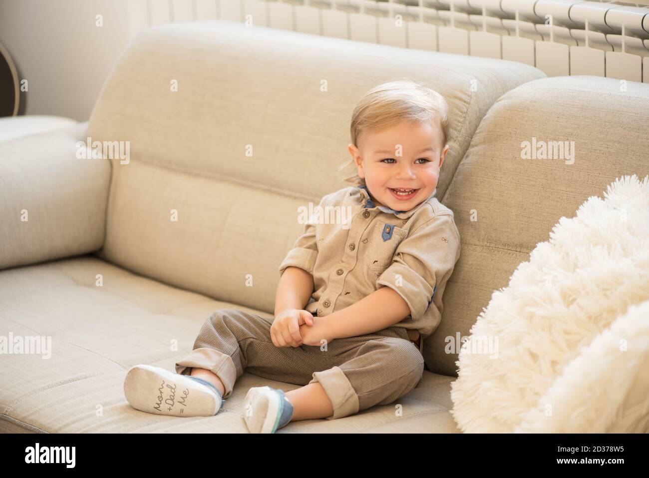 Nicely dressed and happy little boy with blue eyes sitting on the sofa ...