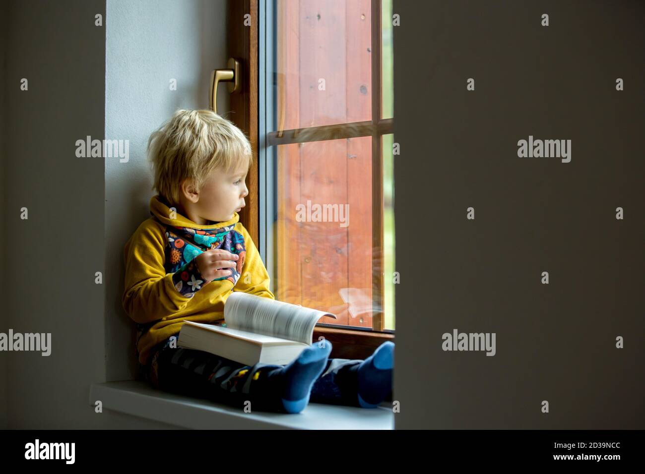 Cute toddler child, blond boy, sitting on window at home, reading book ...