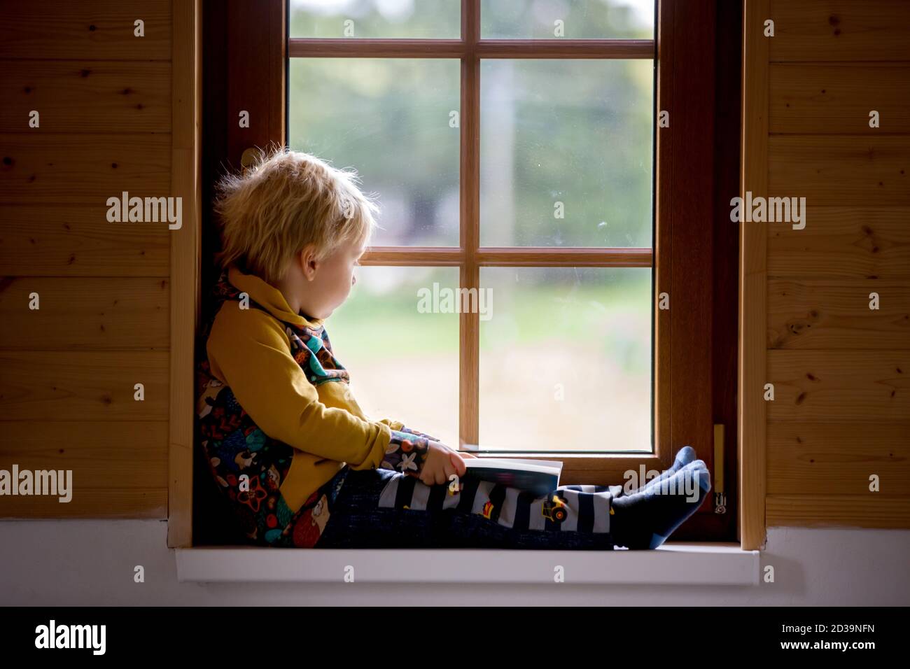 Cute toddler child, blond boy, sitting on window at home, reading book ...
