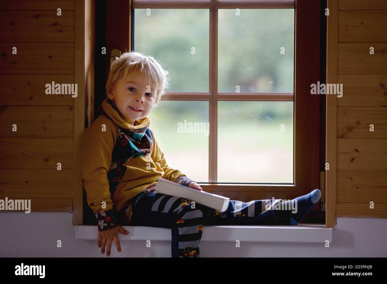 Cute toddler child, blond boy, sitting on window at home, reading book ...