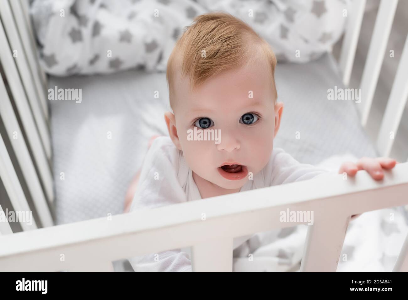 overhead view of baby boy looking at camera while standing in crib with ...