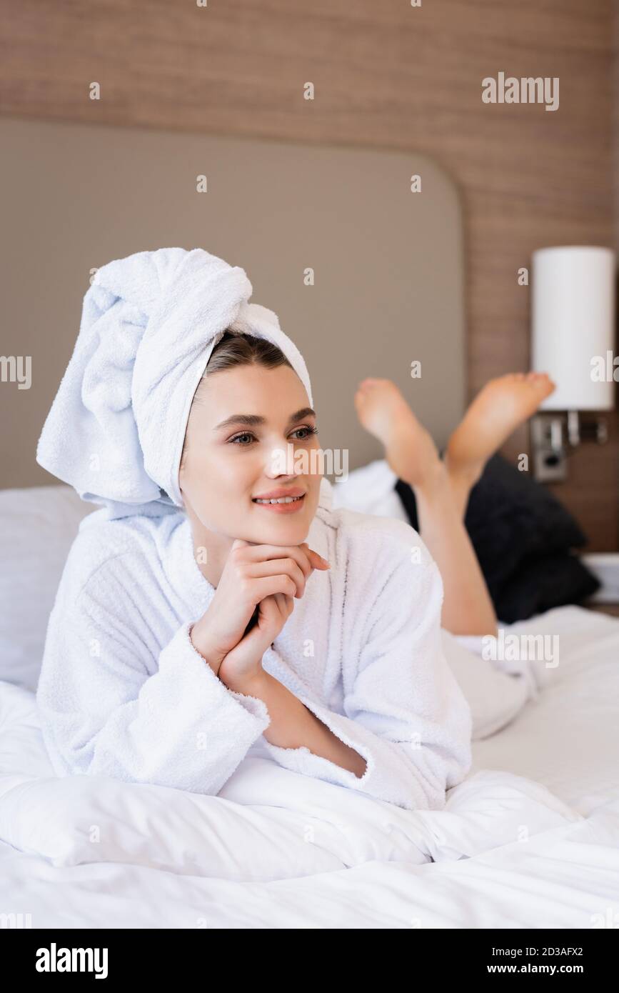 barefoot woman in towel and white bathrobe lying on bed and looking ...