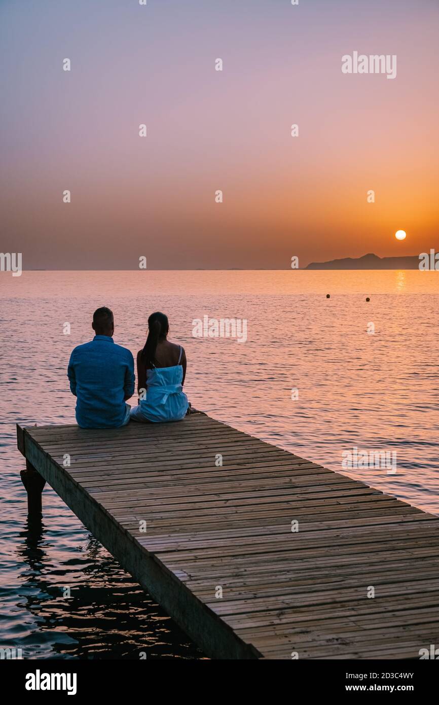 Crete Greece, young romantic couple in love is sitting and hugging on wooden pier at the beach ...