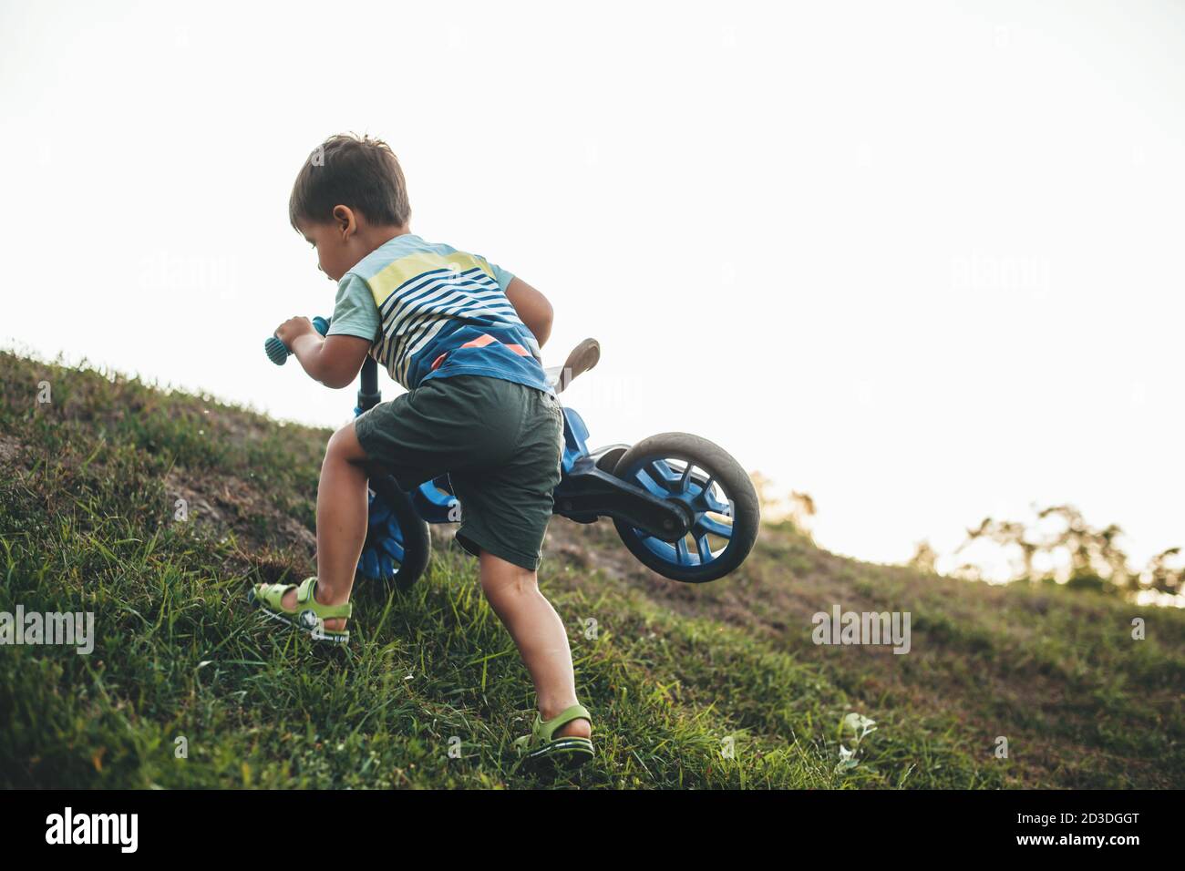 Small caucasian boy pushing his bike on a hill during a walk on a field ...
