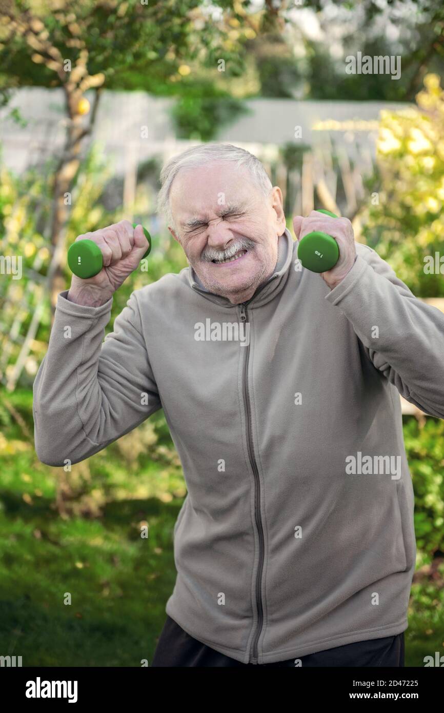 old man trains his muscles with dumbbells with great effort in a Park ...