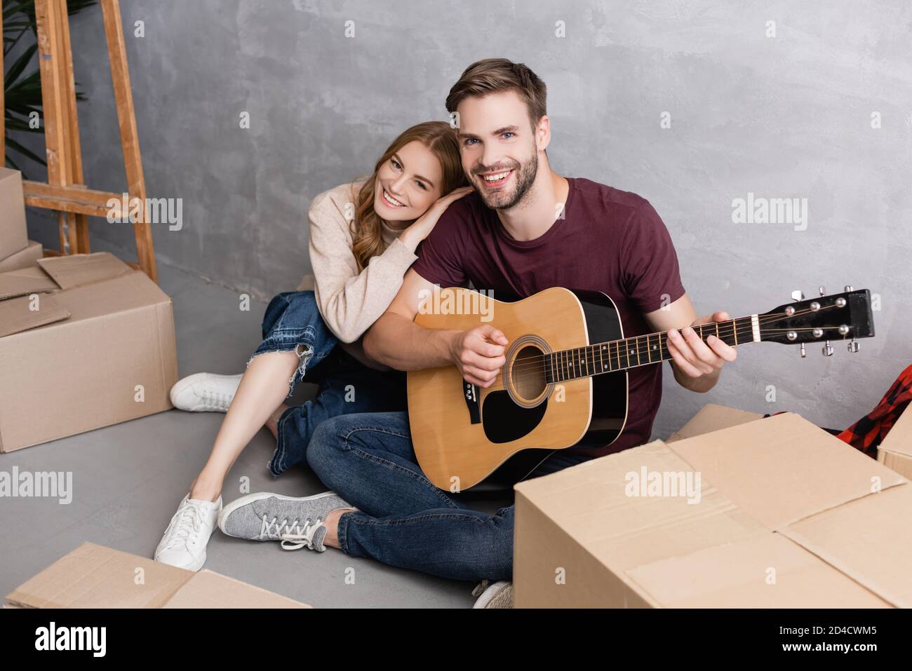pleased woman hugging boyfriend playing acoustic guitar near boxes ...
