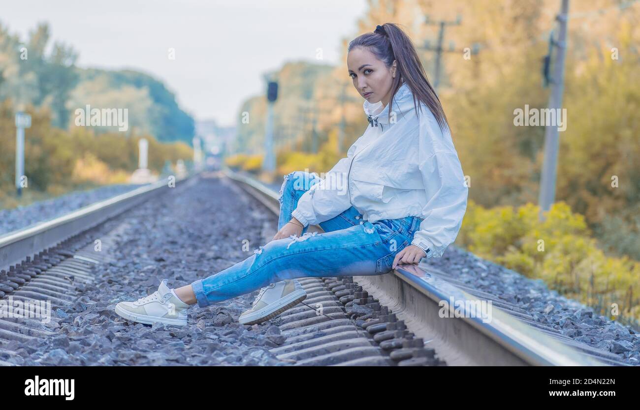 Lonely young woman in a white jacket and jeans sitting on a railway track and looking at the ...