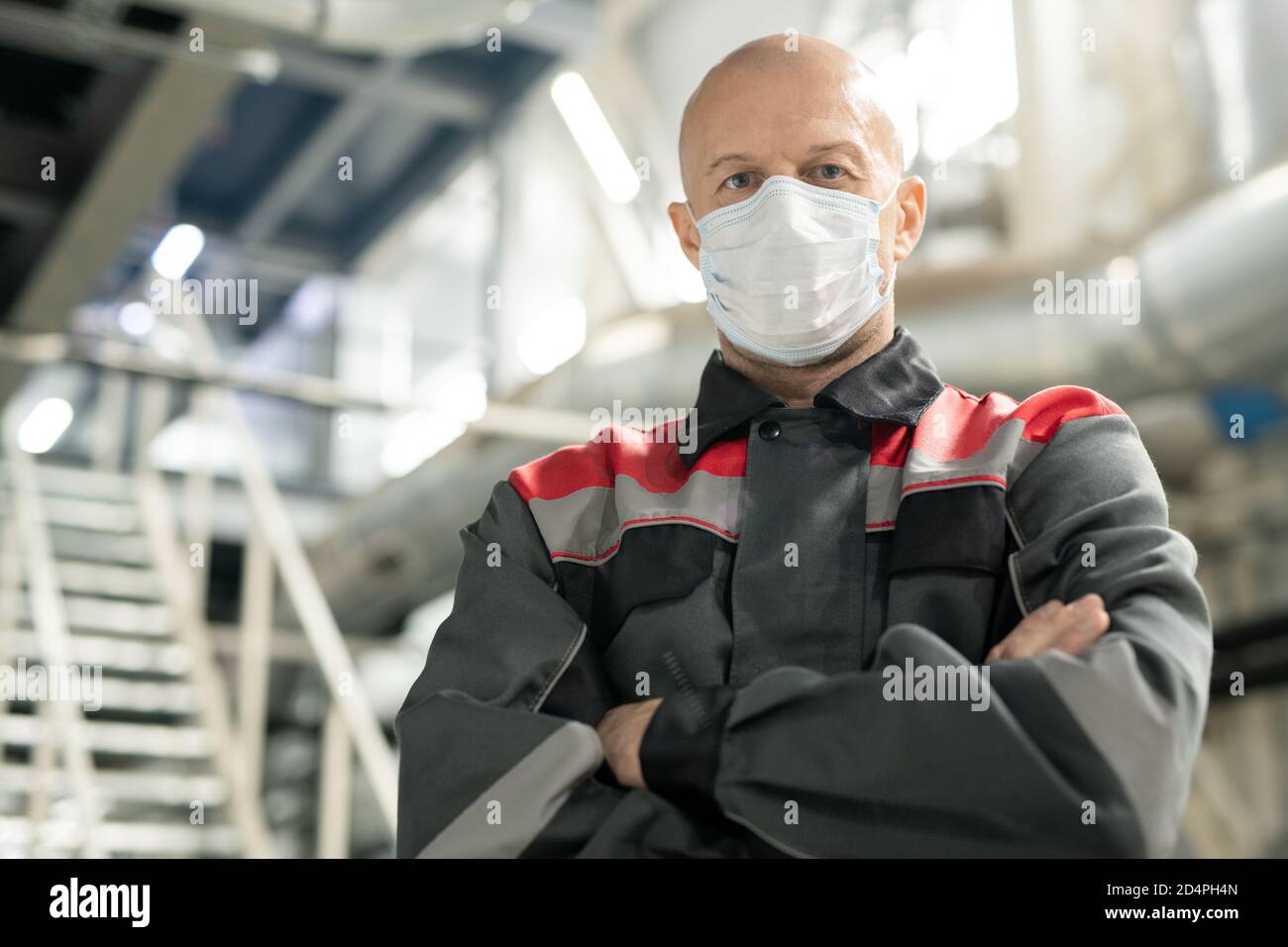 Bald mature worker in uniform and protective mask standing against ...